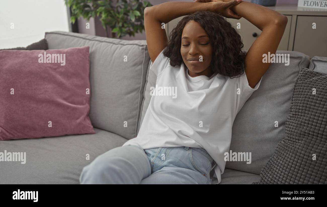 A young african american woman with curly hair relaxes on a couch in a living room, wearing a ...