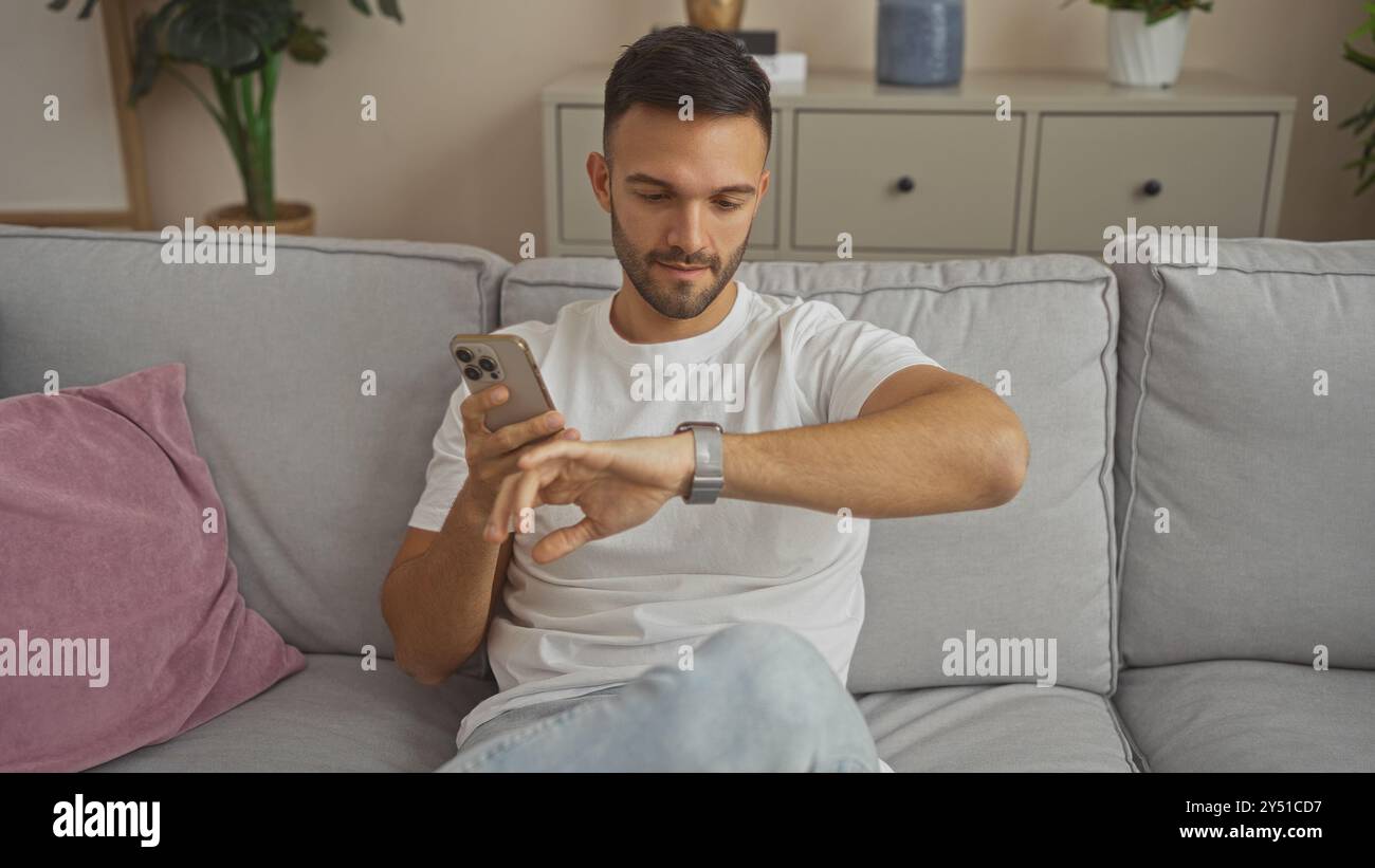Handsome young hispanic man with a beard sitting on a sofa in a living room, checking his ...