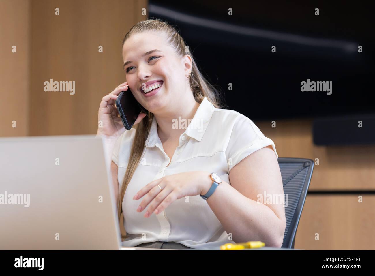 Talking on smartphone, woman smiling while working on laptop in office ...