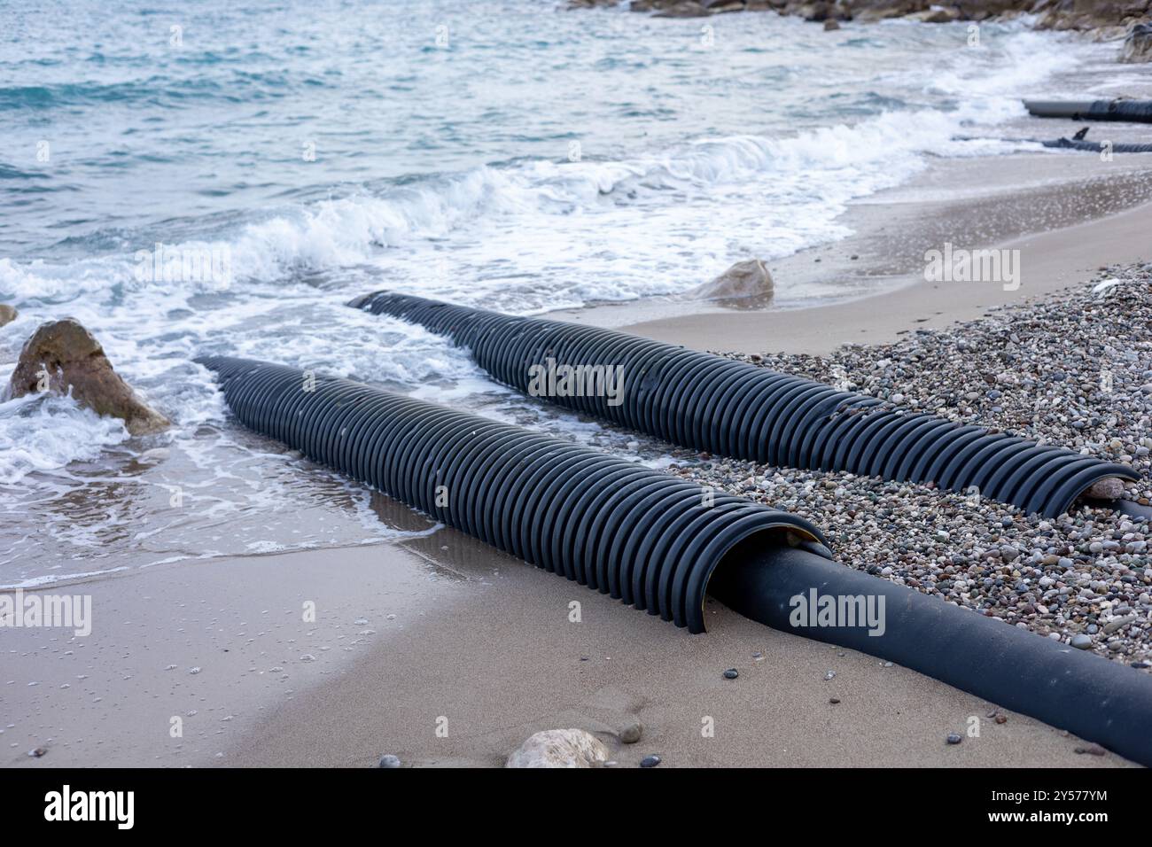 Black drainage pipes washed ashore on a rocky beach with gentle waves ...