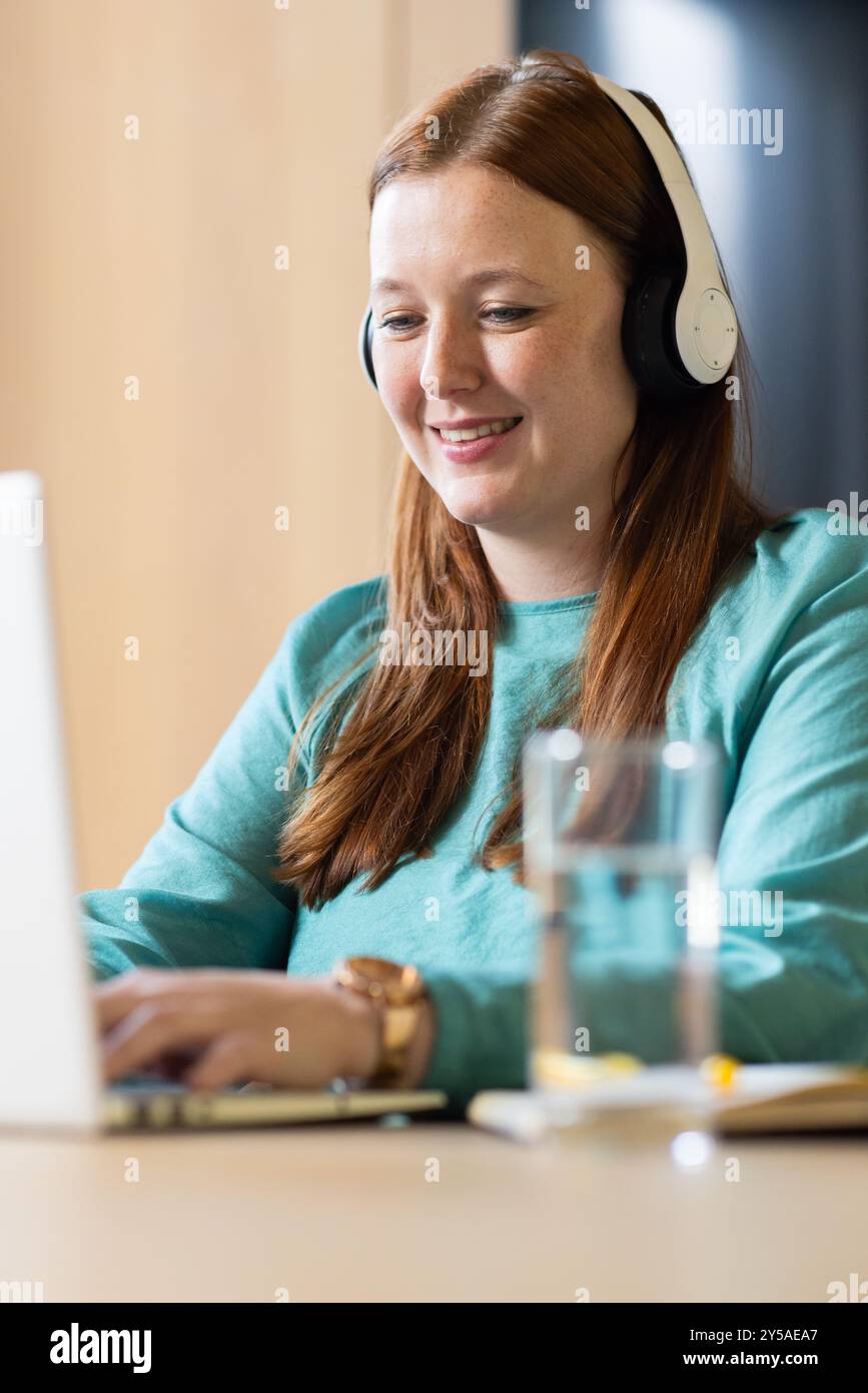 At office, Wearing headphones, woman using laptop and smiling while ...