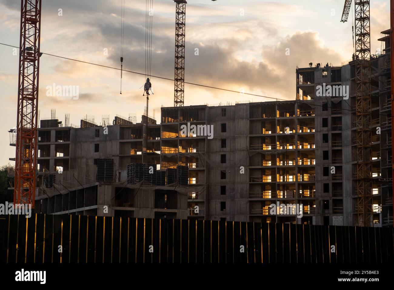 Cranes operate at dusk, lifting materials above a bustling construction ...
