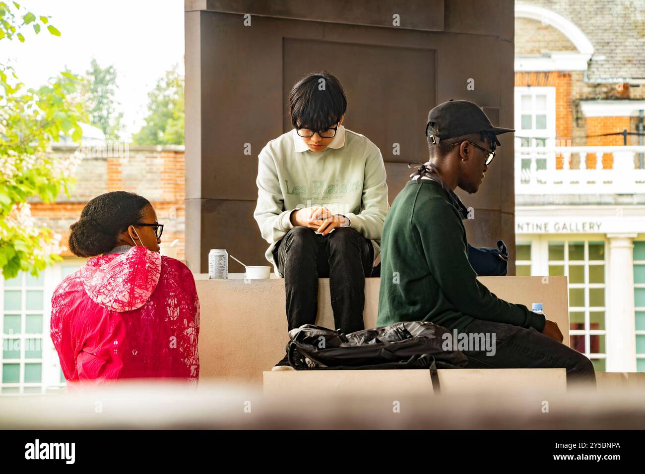 Three young people sit in an outdoor setting, each engaged in their own ...
