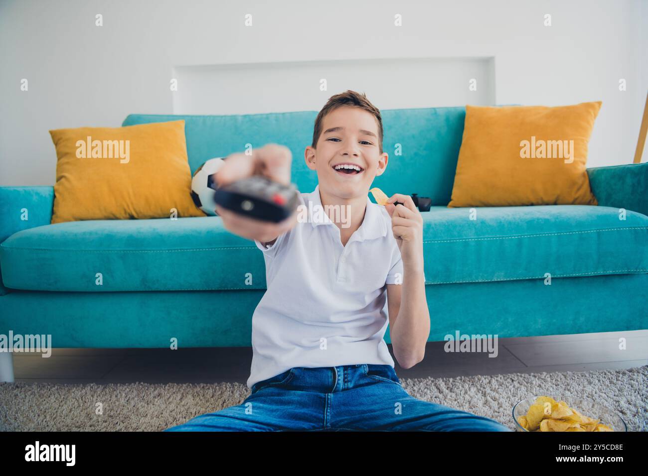 Portrait of charming little boy hold tv remote eat chips wear t-shirt ...