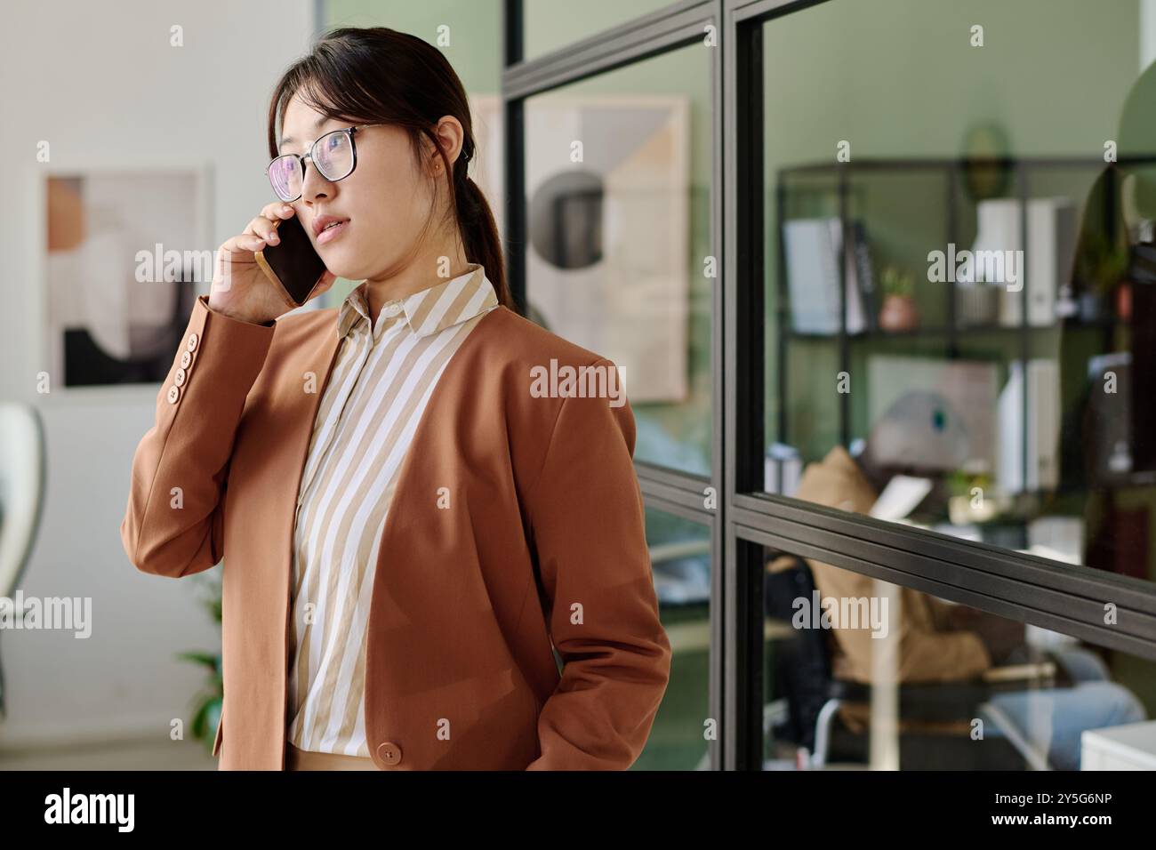 Woman standing in office, holding phone, wearing glasses and blazer ...