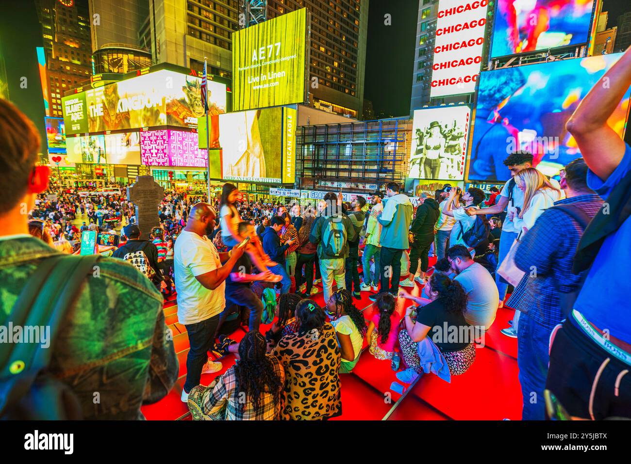People gathered on the red stairs in Times Square at night, surrounded ...