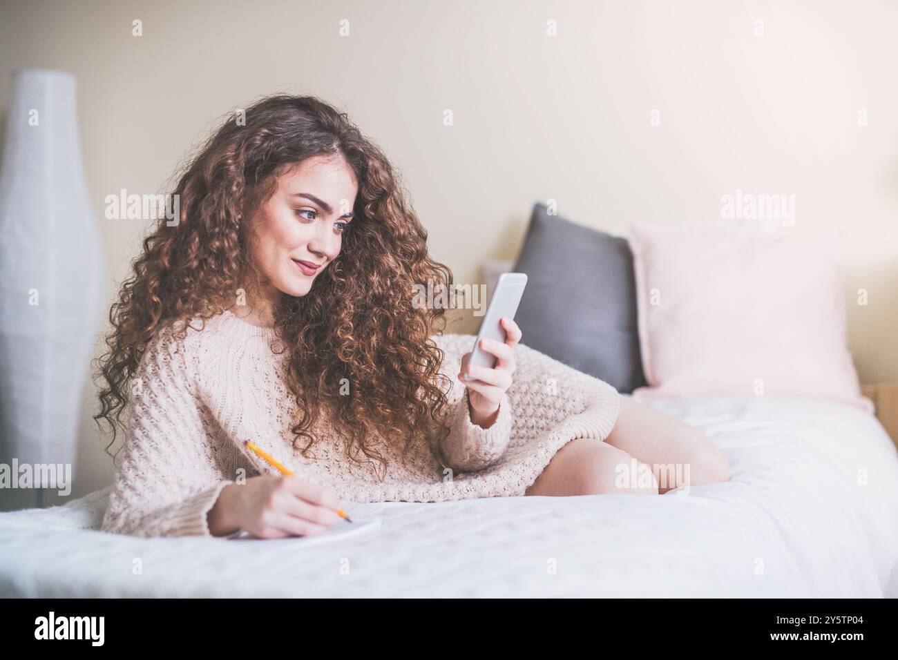 Beautiful young woman lying on her stomach on bed, scrolling on smartphone, smiling Stock Photo ...
