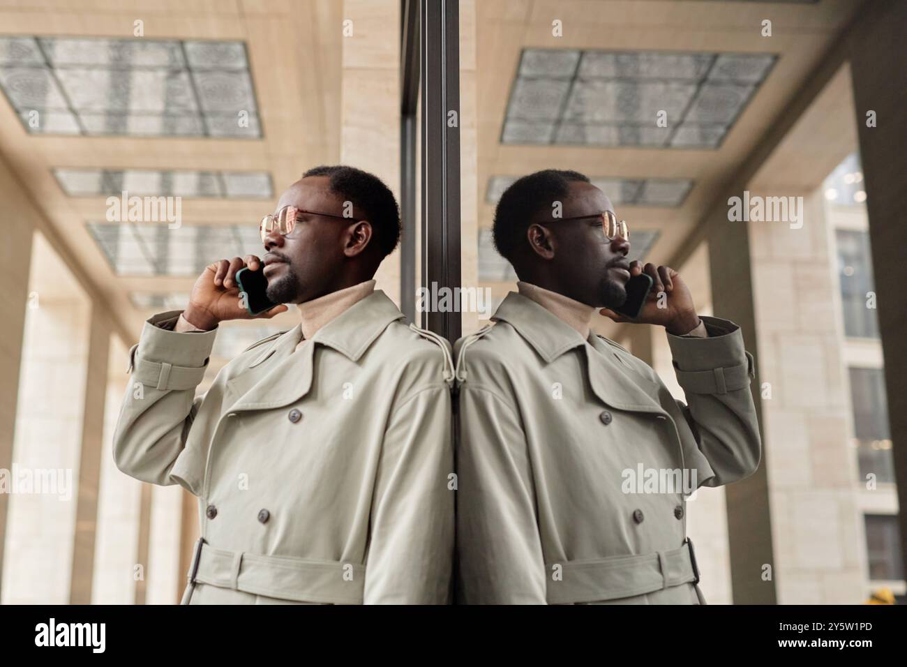 African American man standing next to large window, holding smartphone ...