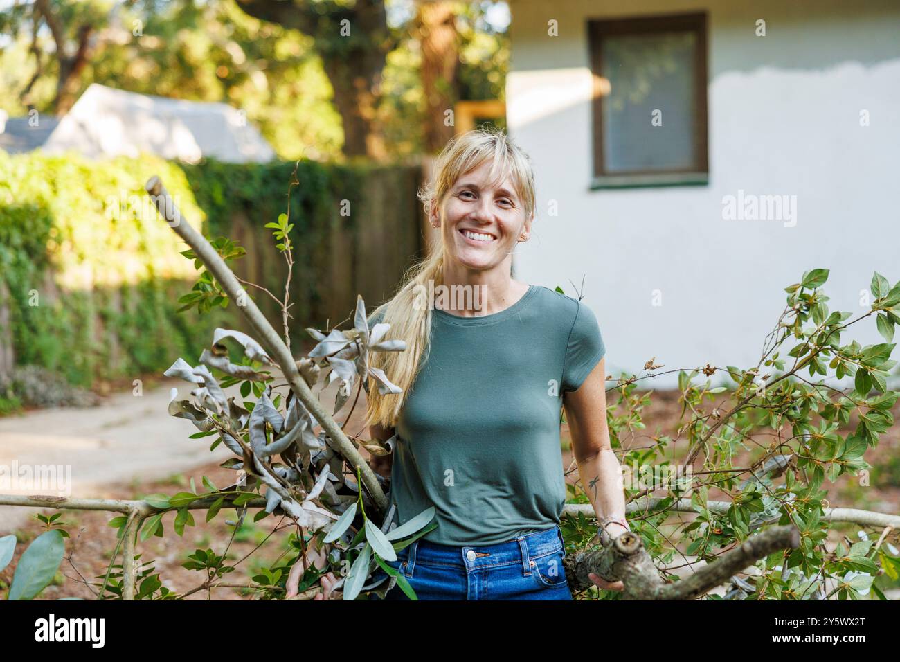 Smiling woman with blond hair holding cut branches while doing yard work outside a house ...