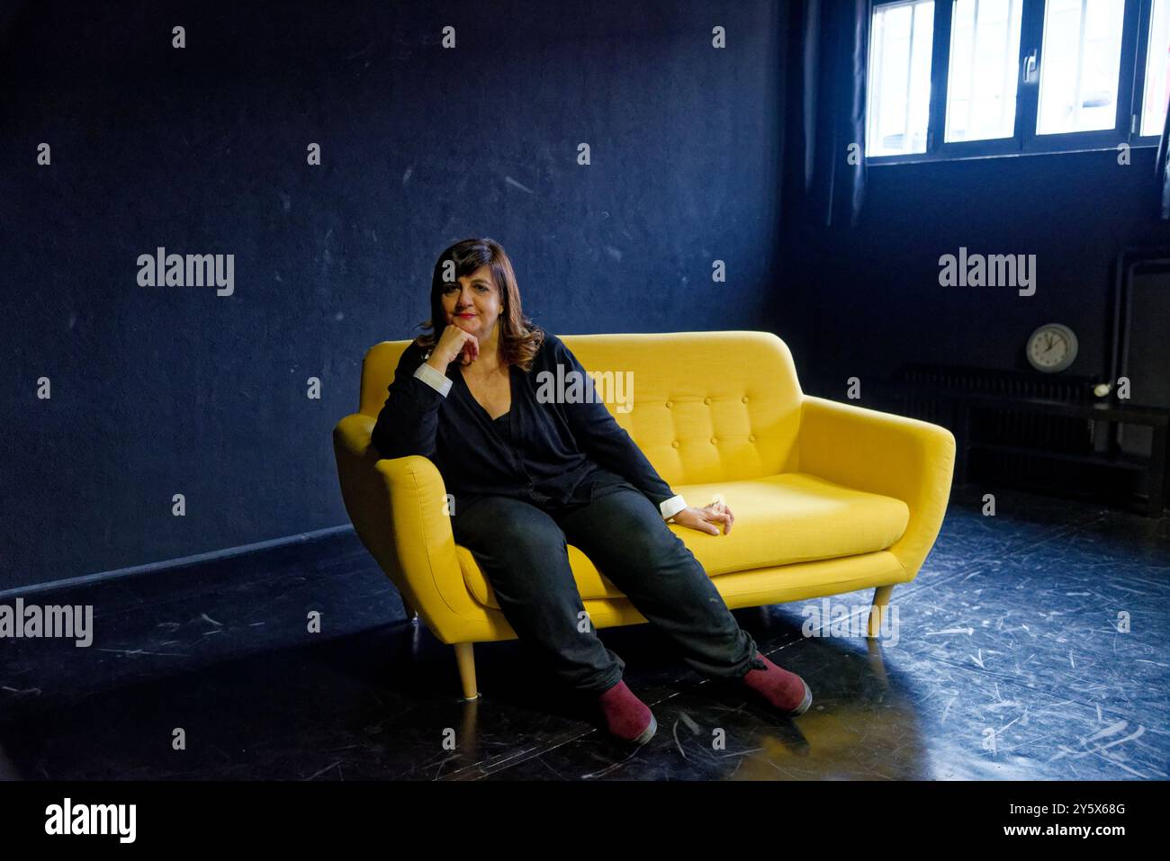 Mature woman seated thoughtfully on a yellow sofa in a dimly lit room ...