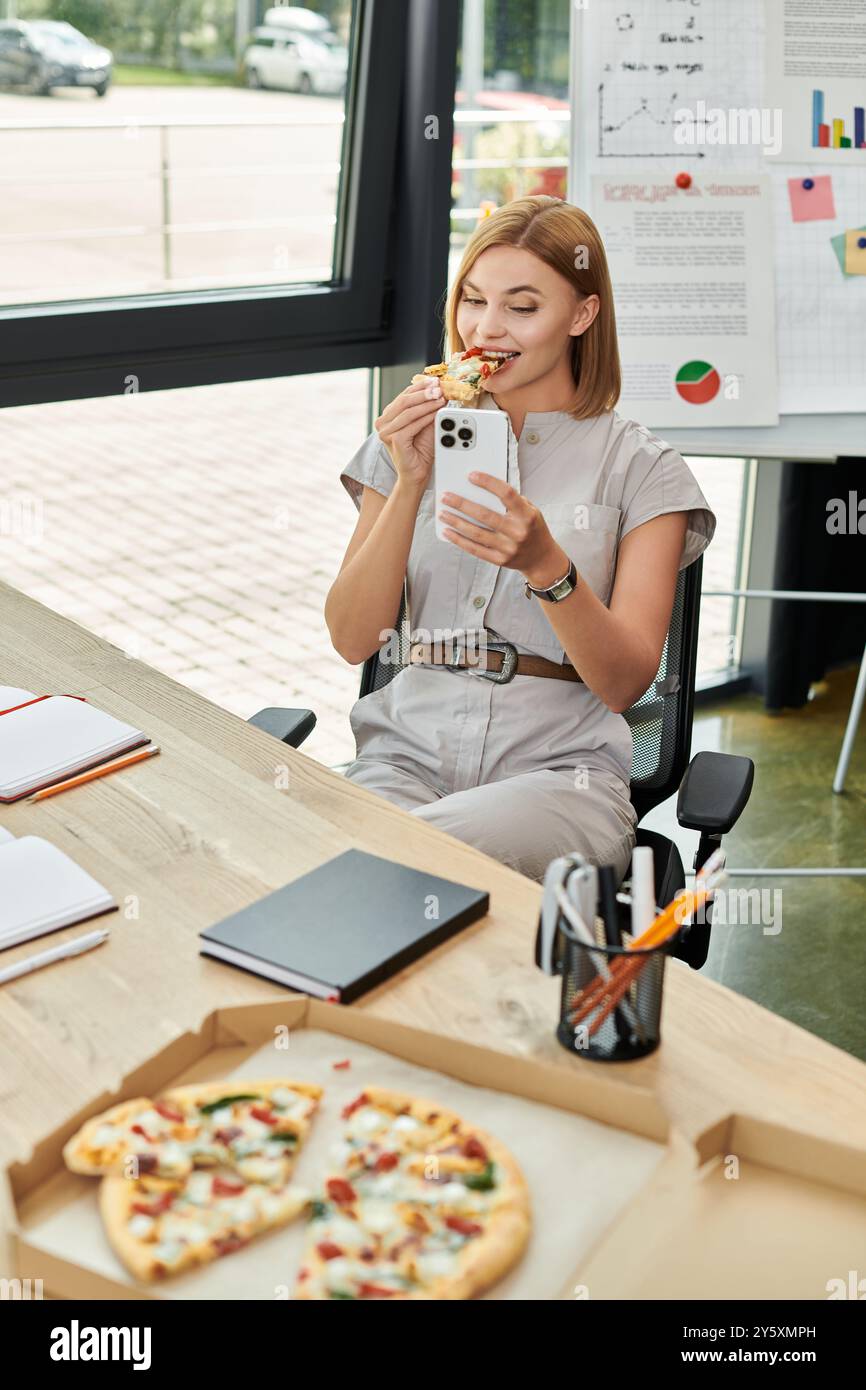 A young woman takes a bite of pizza while checking her smartphone at ...
