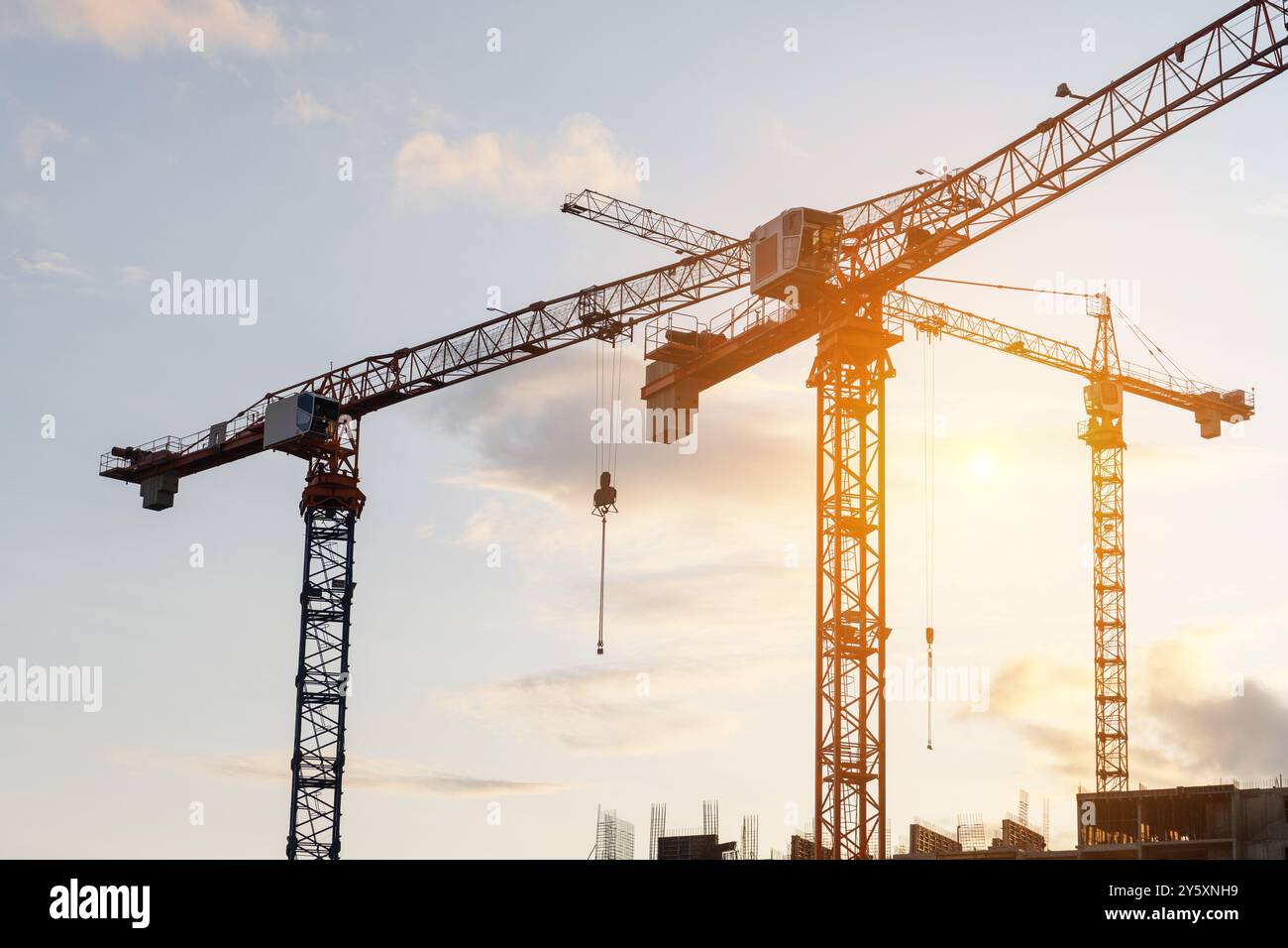 Cranes are actively lifting materials at a construction site as the sun sets, highlighting the ...