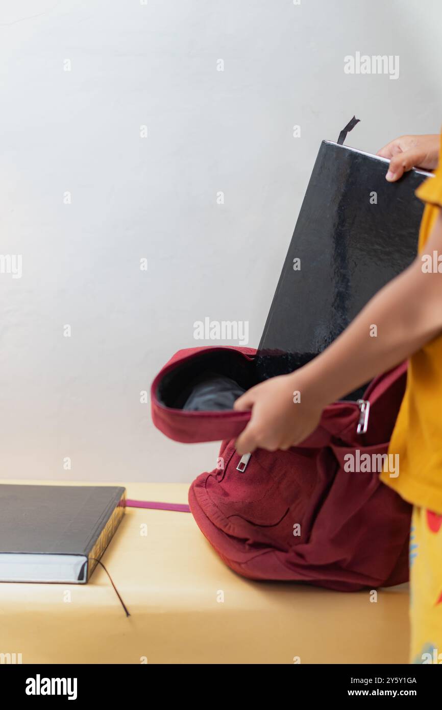 A child packs their backpack with large books for school. The red bag ...