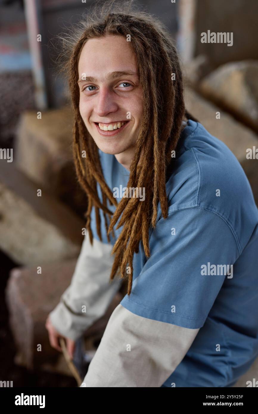 Young man with dreadlocks smiling while sitting on rocky surface and ...