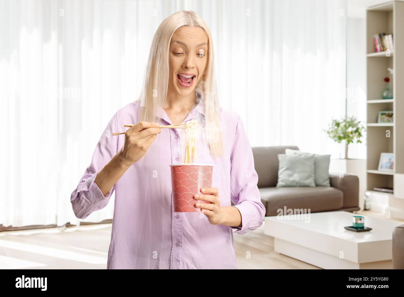 Excited young woman eating noodles from a takeaway box at home in a ...