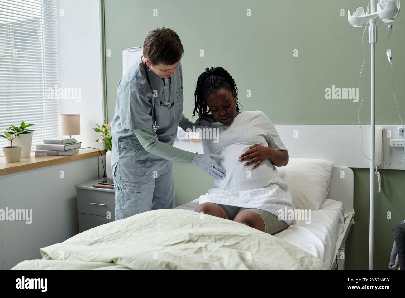Medical professional assisting pregnant woman in hospital room, providing care and support ...