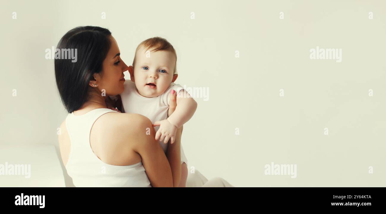 Happy young mother holding her cute baby sitting together in white room at home, studio ...