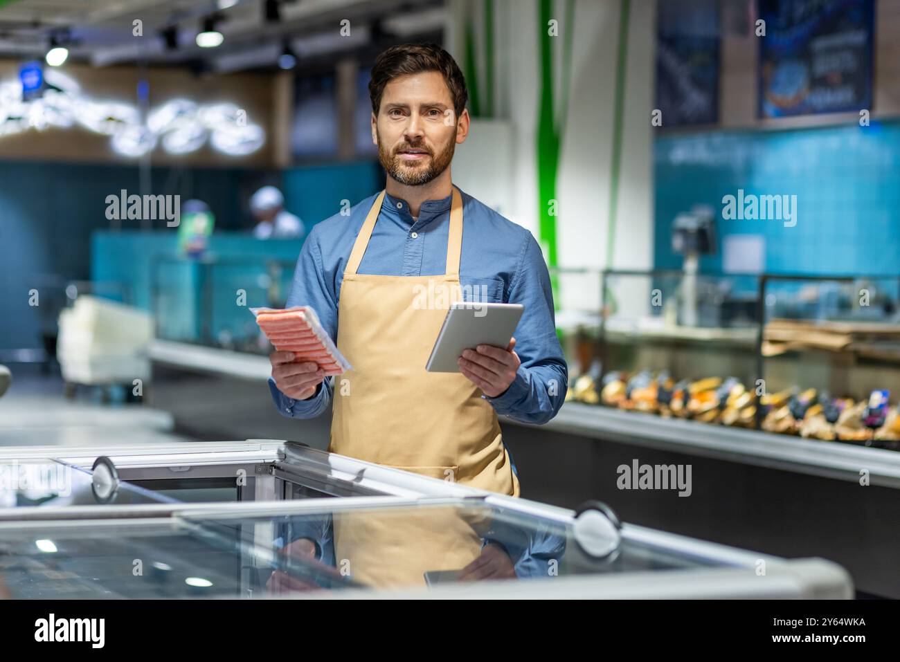 Clerk wearing apron stands in supermarket holding tablet and packaged ...