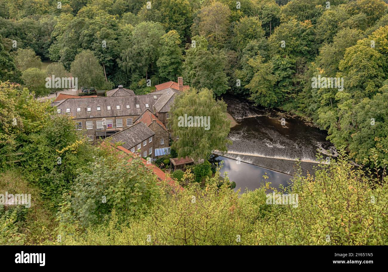 Aerial view of buildings around a mill and a weir. A woodland of trees ...
