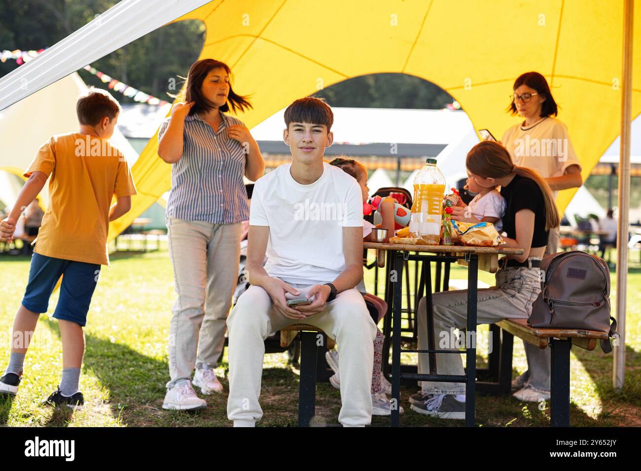 A family group relaxes outdoors under a yellow tent, sharing food and ...