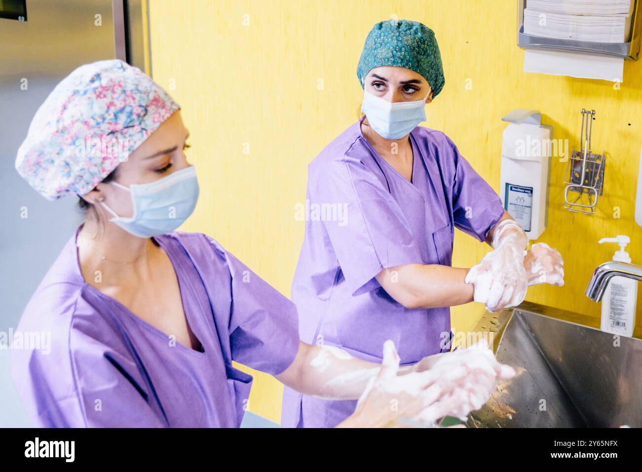 Two female healthcare workers in purple scrubs and surgical caps wash ...