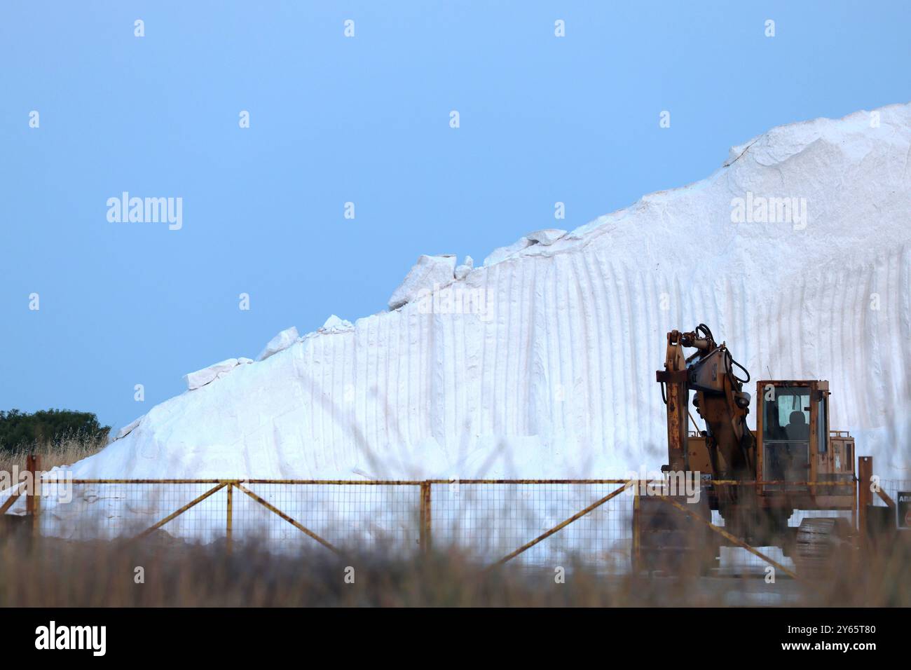 Excavator operating in front of a large salt pile with a clear blue sky ...