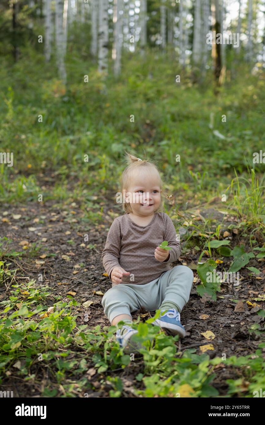 A happy toddler sits on the forest ground, surrounded by lush greenery ...