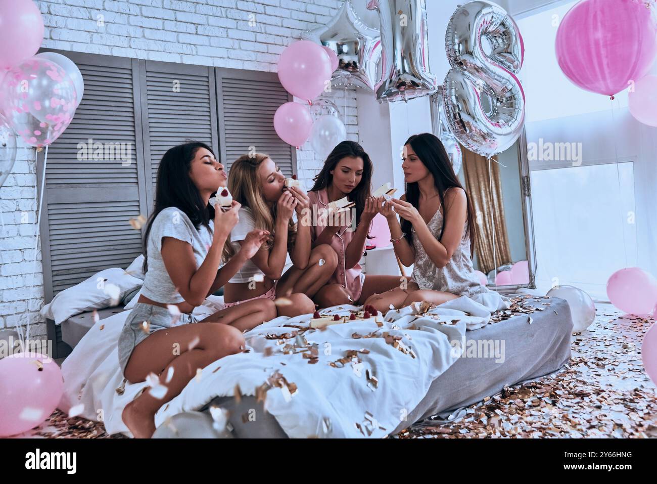 Hungry girls. Four beautiful young women in pajamas eating cake while having a slumber party in ...