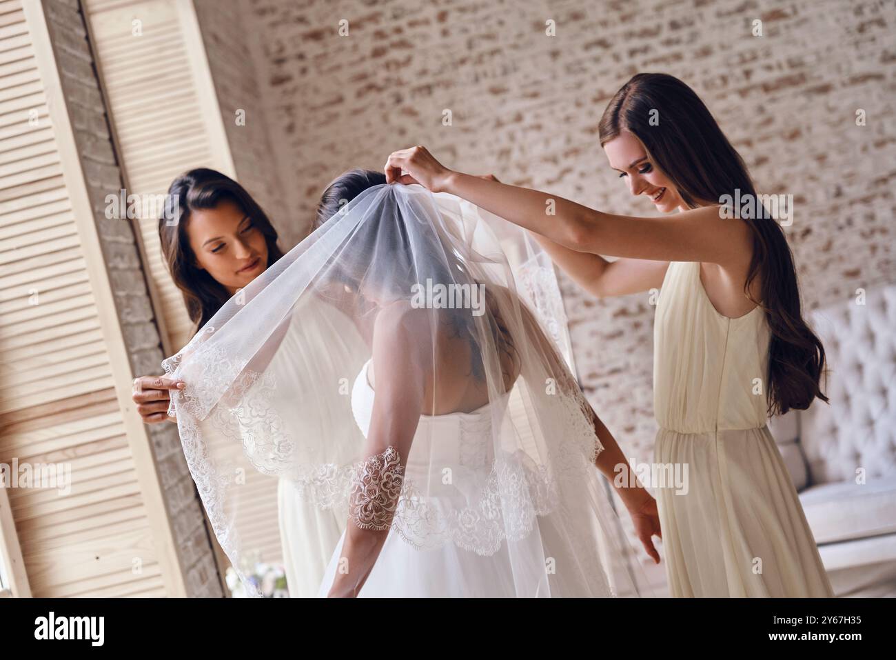 Last preparations. Bridesmaids helping bride to put on a veil while ...