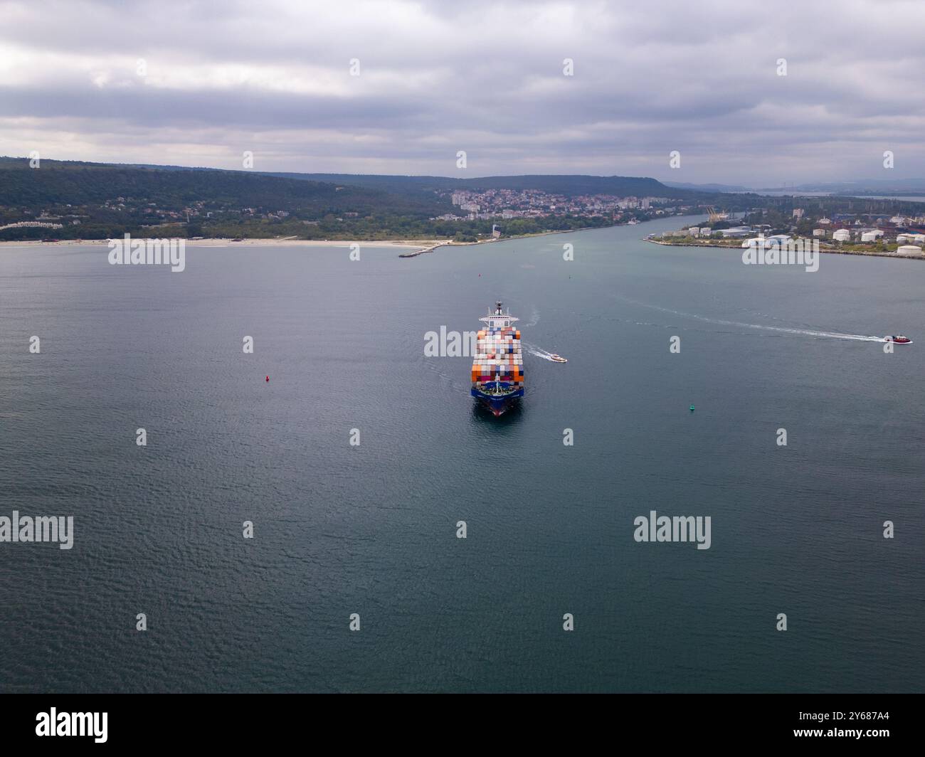 A large cargo container ship moves through the harbor waters, flanked ...