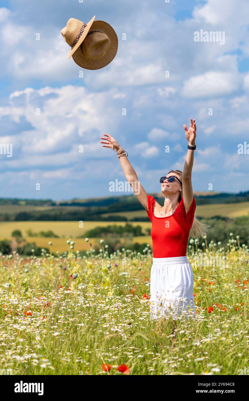 Blonde woman in sunglasses in a wildflower field throwing straw hats in ...