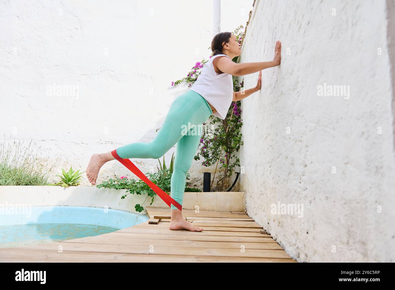 Woman performs leg exercises with a red resistance band by the pool ...