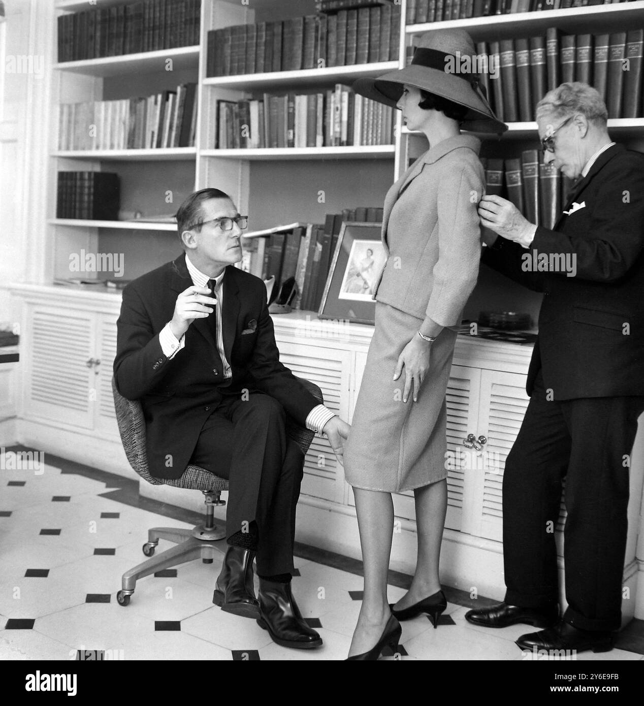 7 DECEMBER 1962 HARDY AMIES, WEARING CHELSEA BOOTS, EXAMINES AN OUTFIT ...