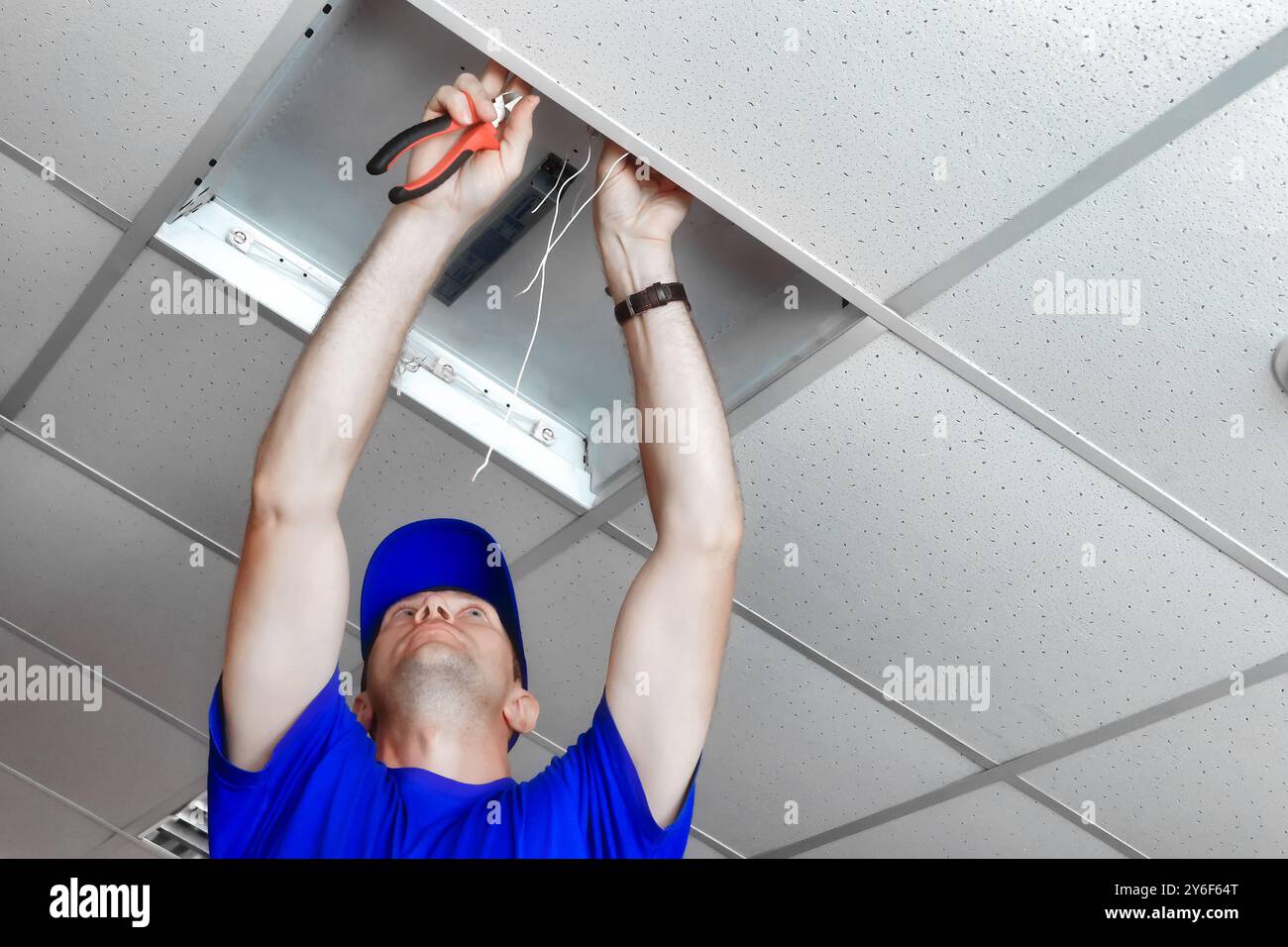 This image depicts an electrician dressed in blue working on ceiling ...