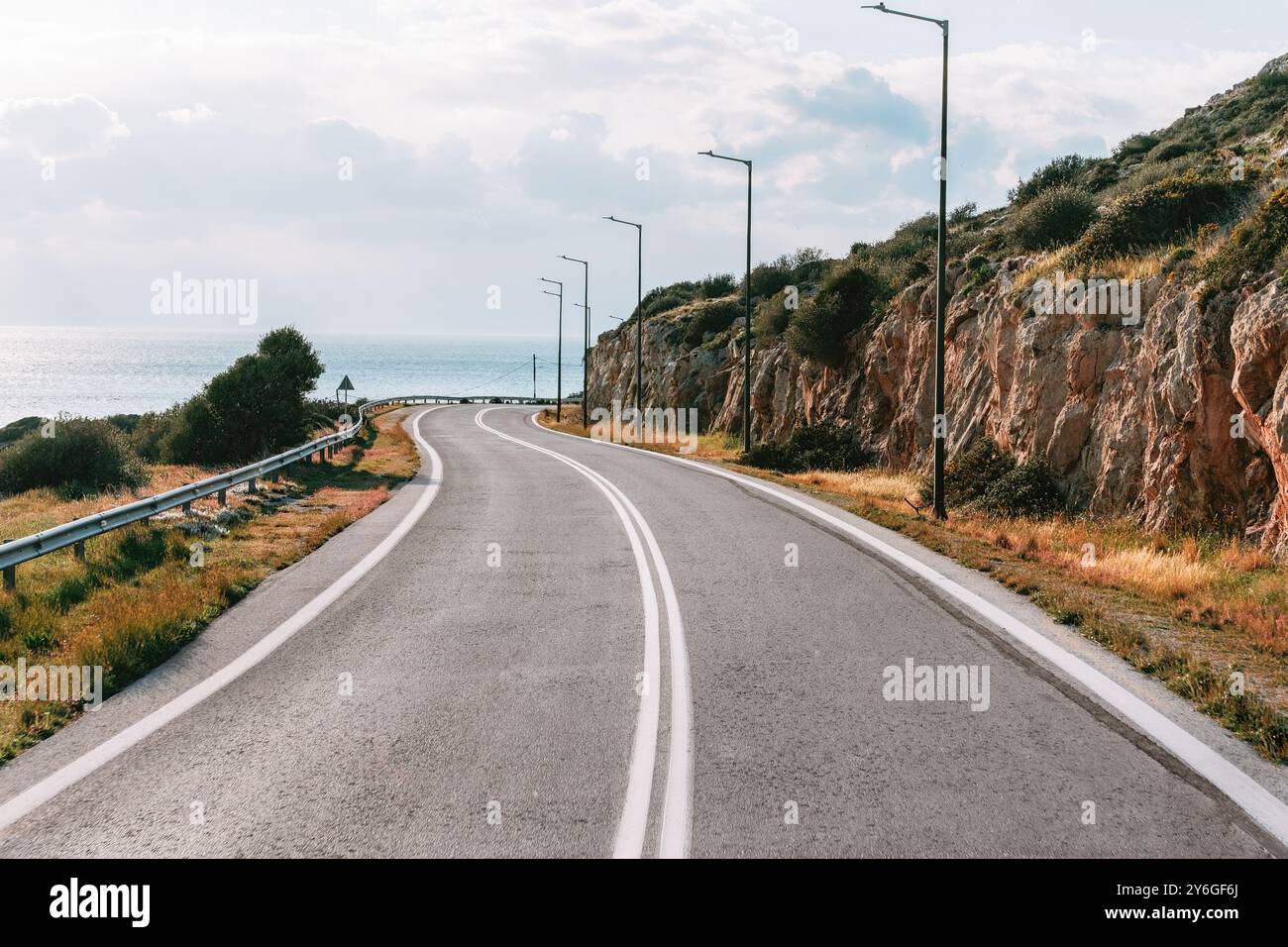 Empty curved road with road markings runs by rocky cliffs with the sea ...
