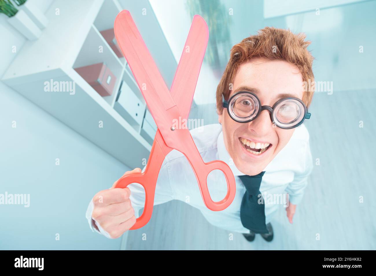 Smiling businessman in an office, holding giant red scissors, exudes ...