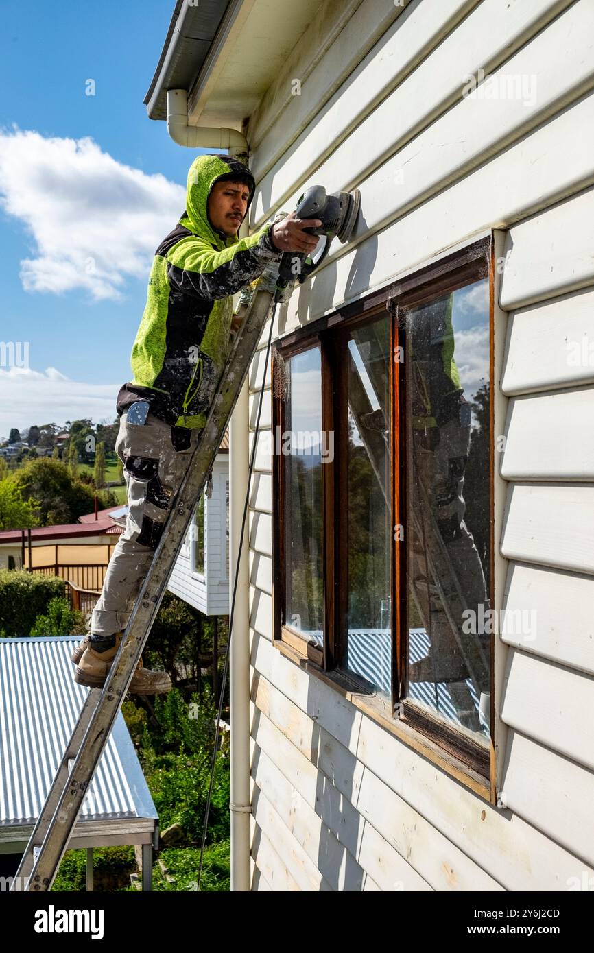 Painter on ladder doing surface preparation on a timber weatherboard ...