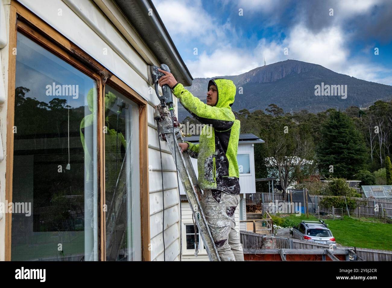 Painter on ladder doing surface preparation on a timber weatherboard ...