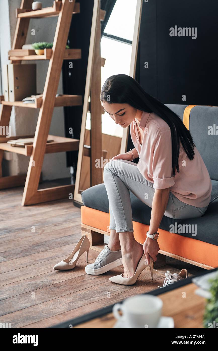Perfect shoes. Thoughtful young woman trying on elegant shoes with high heels while sitting in ...