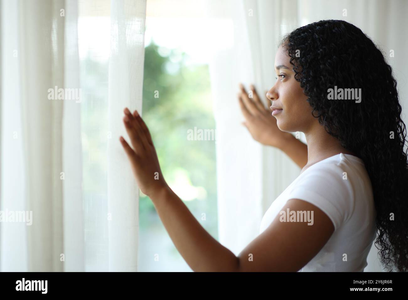 Serious black woman opening curtains and looking through a window at ...