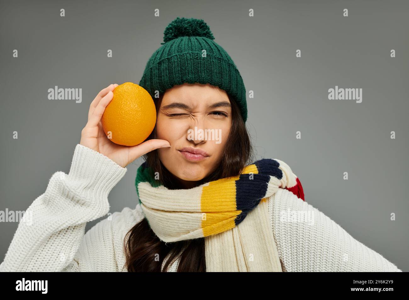Expressing joy, a stylish young woman winks while holding an orange ...