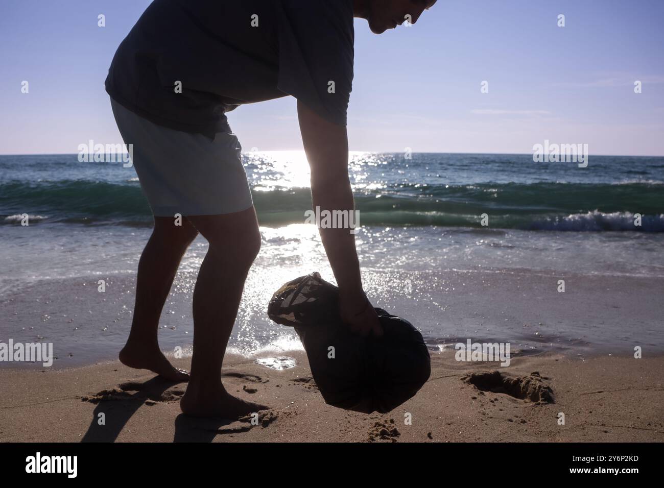 Young man picks up a black plastic bag of garbage on the beach barefoot ...