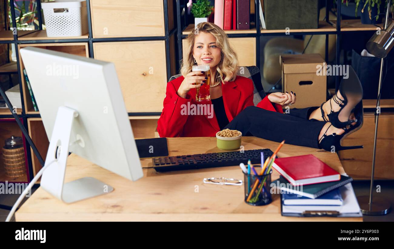 Kicking back with beer, feet up on desk, woman enjoys little downtime ...