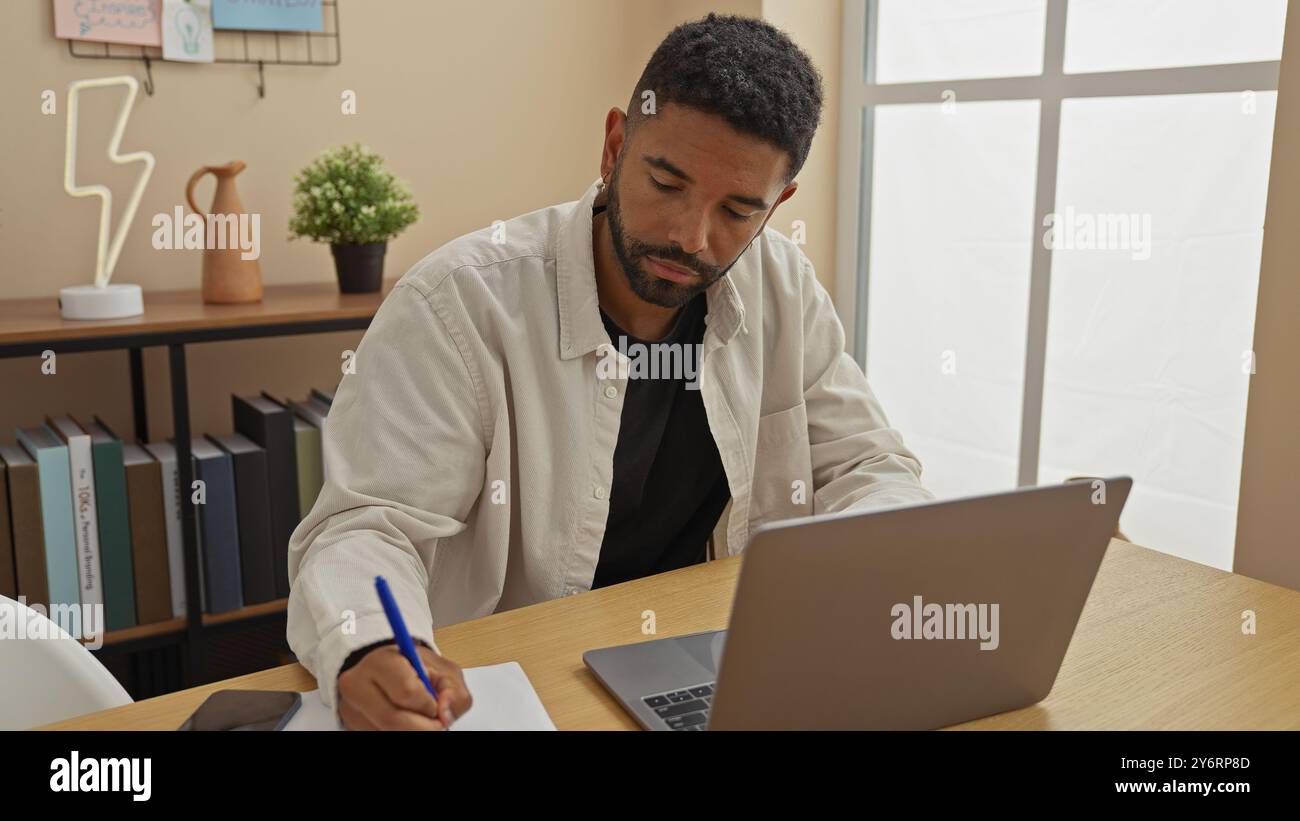 A focused young black man with a beard writing notes and using a laptop ...