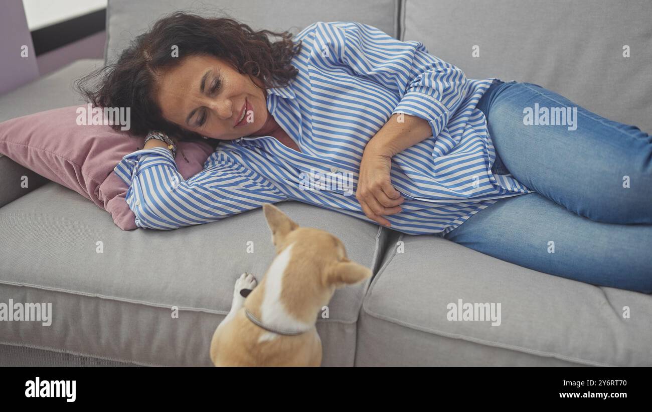 A middle-aged hispanic woman relaxes at home on the couch, enjoying time with her pet chihuahua ...