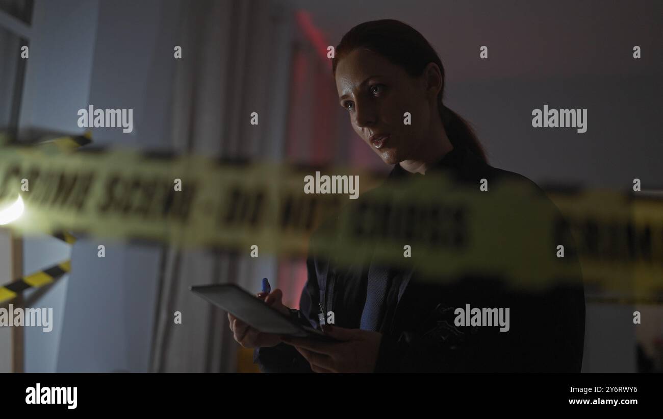 A serious young woman takes notes at an indoor crime scene with yellow ...