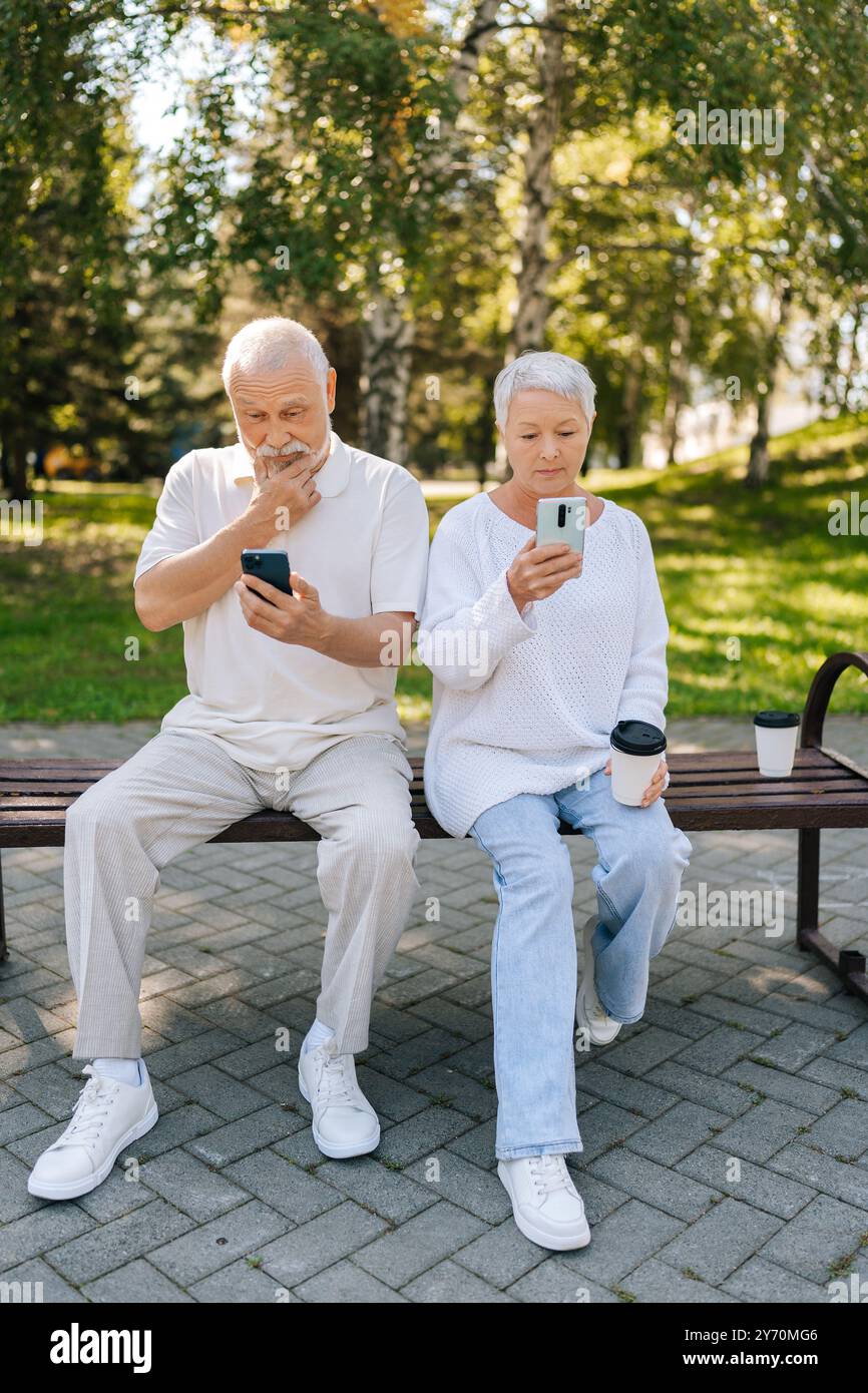 Vertical portrait of beautiful elderly couple joyfully using smartphones sitting on park bench ...