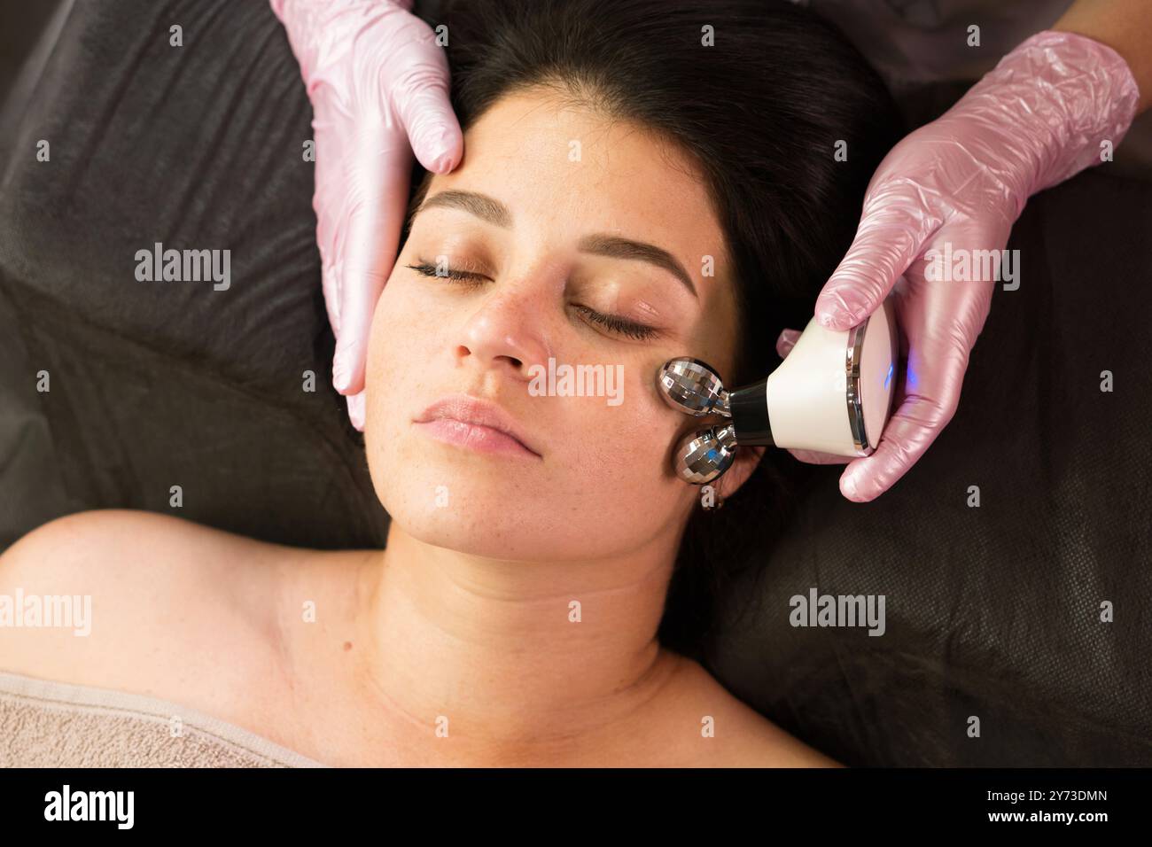 A beautician uses a microcurrent device on a client's cheek, performing ...