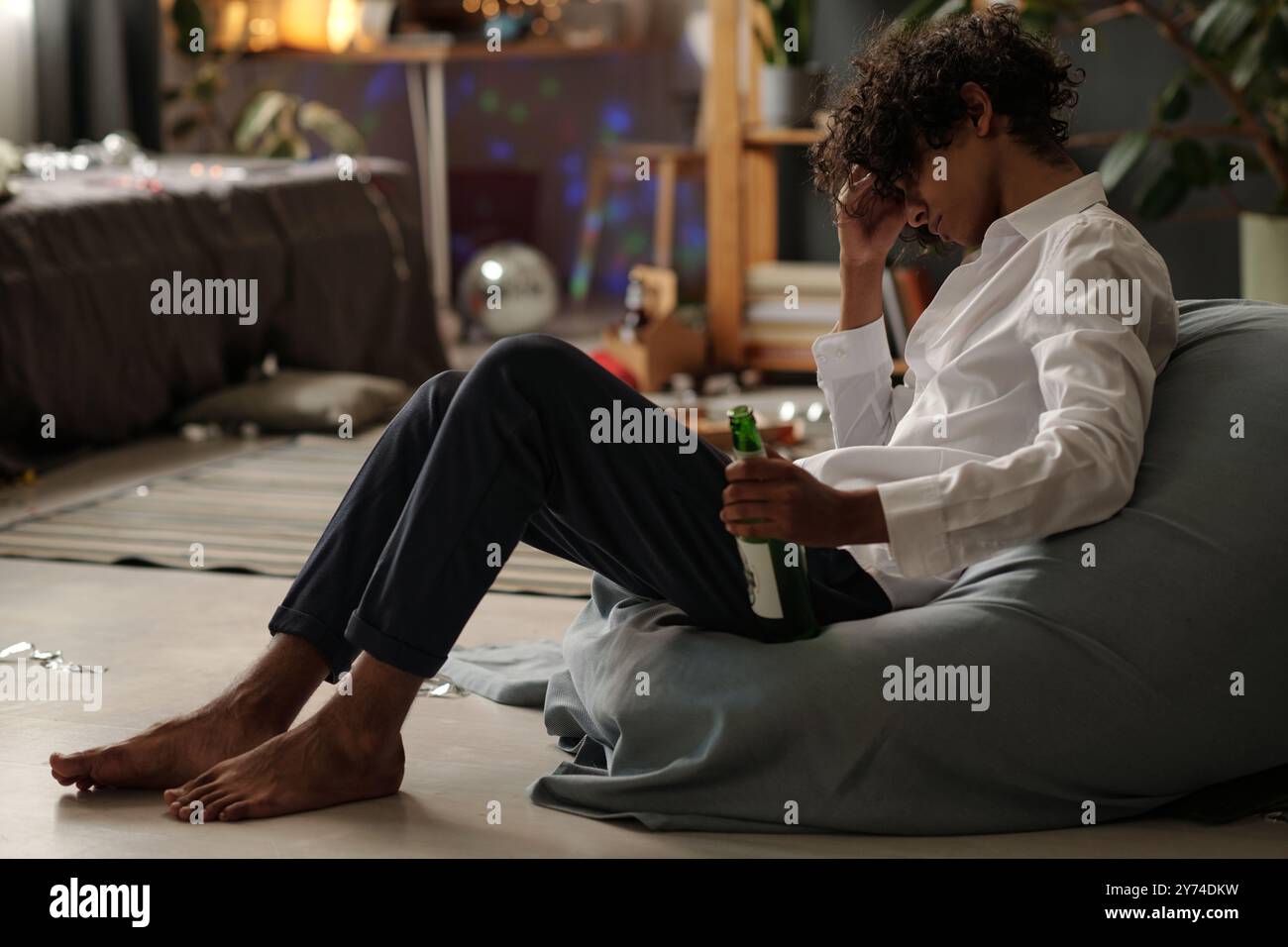 Side view of teenage guy with bottle of beer sitting in sack armchair ...