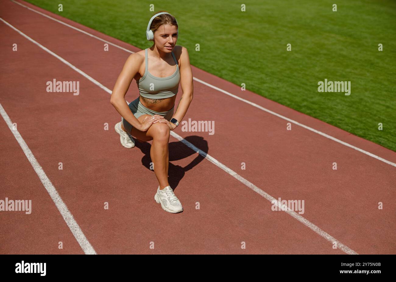A fit woman stretching on a running track, wearing headphones and ...